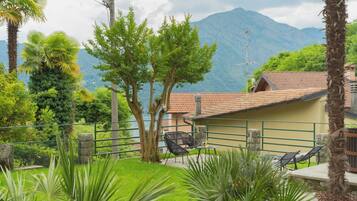 Uma casa com telhado de telhas, jardim cercado e montanha ao fundo.