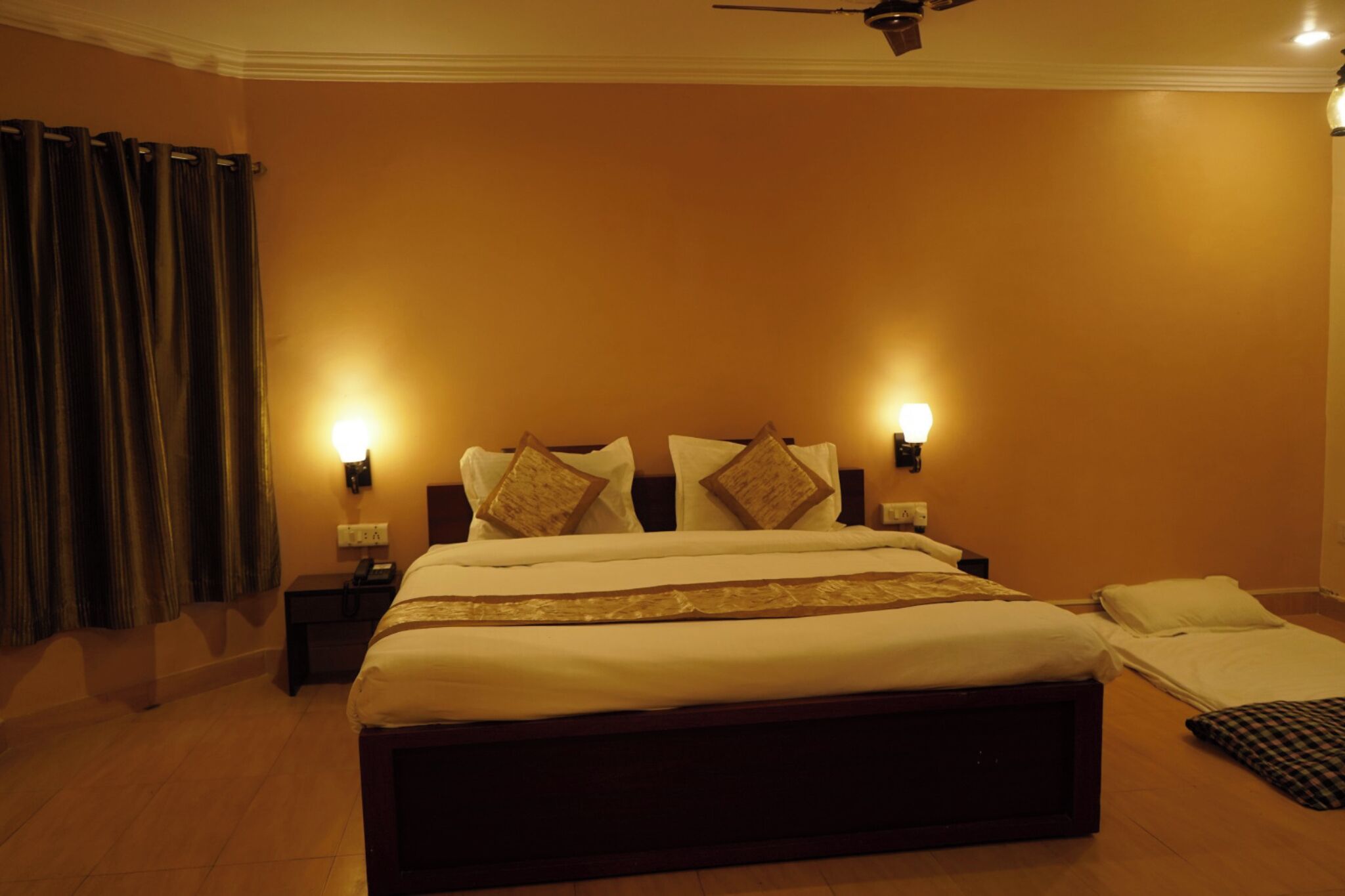 Quarto de hotel com uma cama grande, duas mesas de cabeceira, um criado-mudo, um ventilador de teto e uma janela com cortinas.