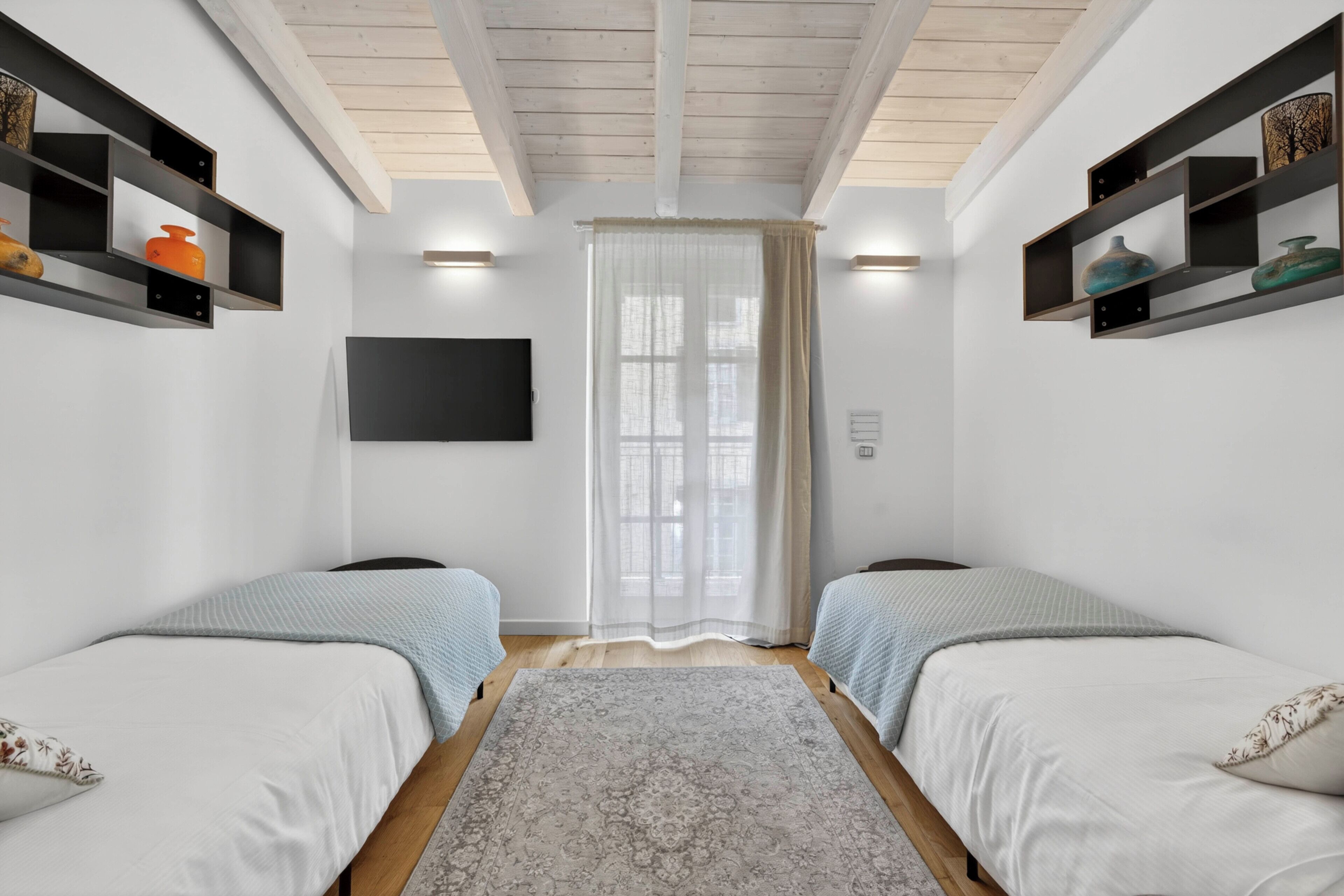Quarto com duas camas, televisão de tela plana e prateleiras com objetos decorativos.