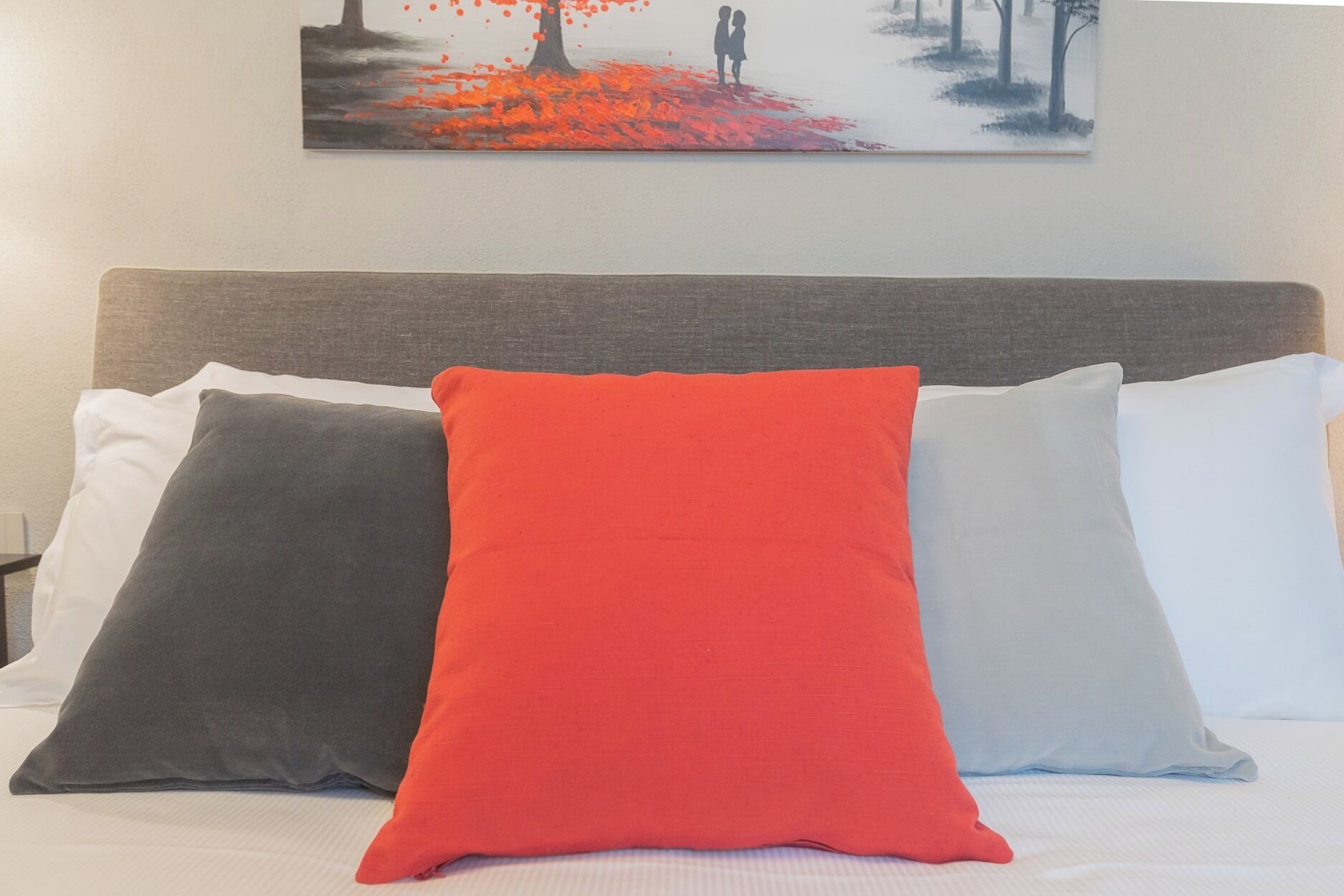 Uma cama com três almofadas de cores diferentes e um quadro em cima.