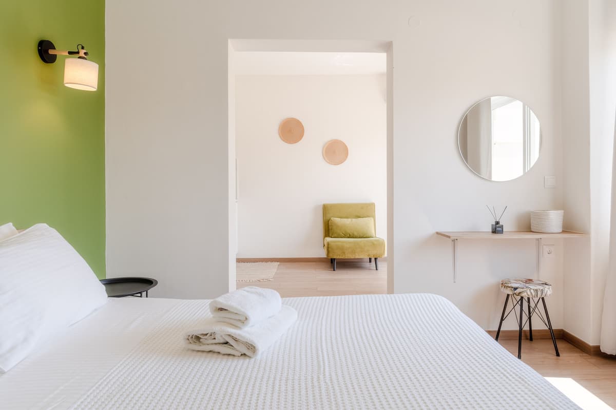 Quarto de hotel com cama, cadeira, um banquinho pequeno, espelho e prateleira com cesta.