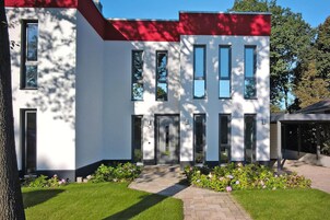 Um edifício moderno de dois andares, com telhado vermelho, janelas amplas e um jardim bem cuidado com flores rosa.