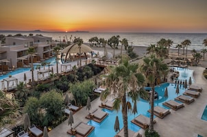 Resort com várias piscinas, palmeiras e vista para o pôr do sol sobre o oceano.