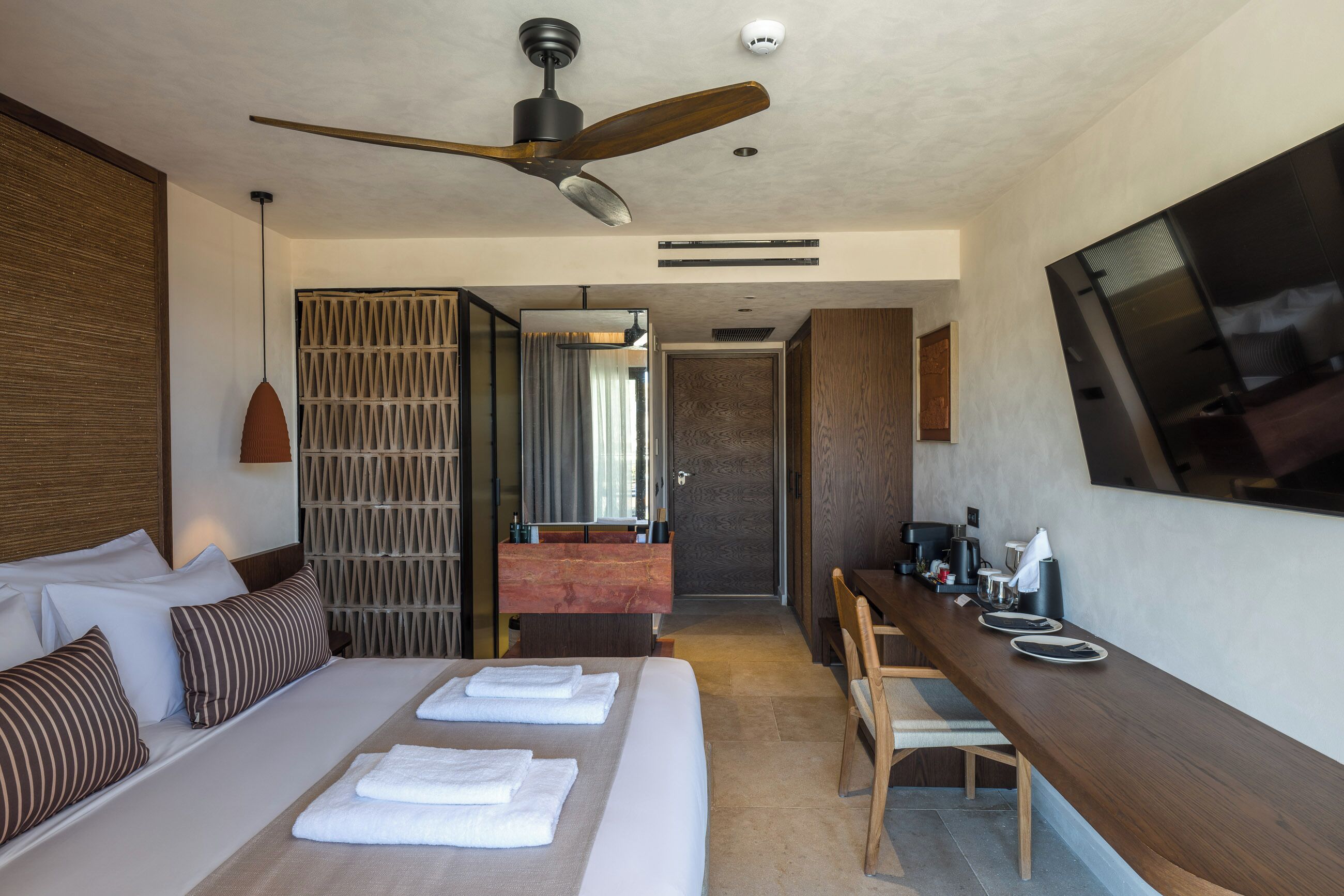 Quarto de hotel com cama, escrivaninha, espelho e ventoinha no teto.