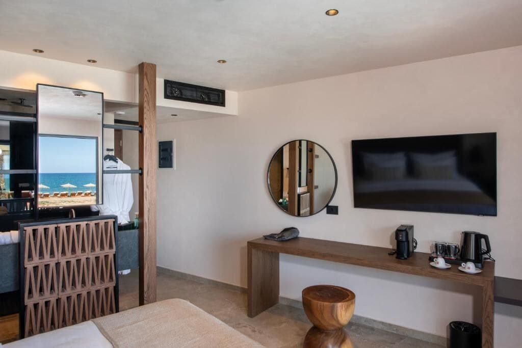 Quarto de hotel moderno com uma cama, uma mesa de console de madeira, um espelho redondo, uma televisão e vista para a praia.