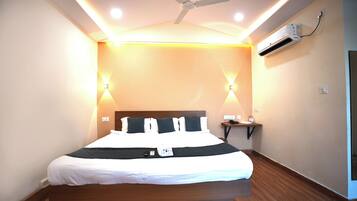 Quarto de hotel com cama, ventilador de teto, ar condicionado fixado na parede, luminárias laterais e mesa de cabeceira.