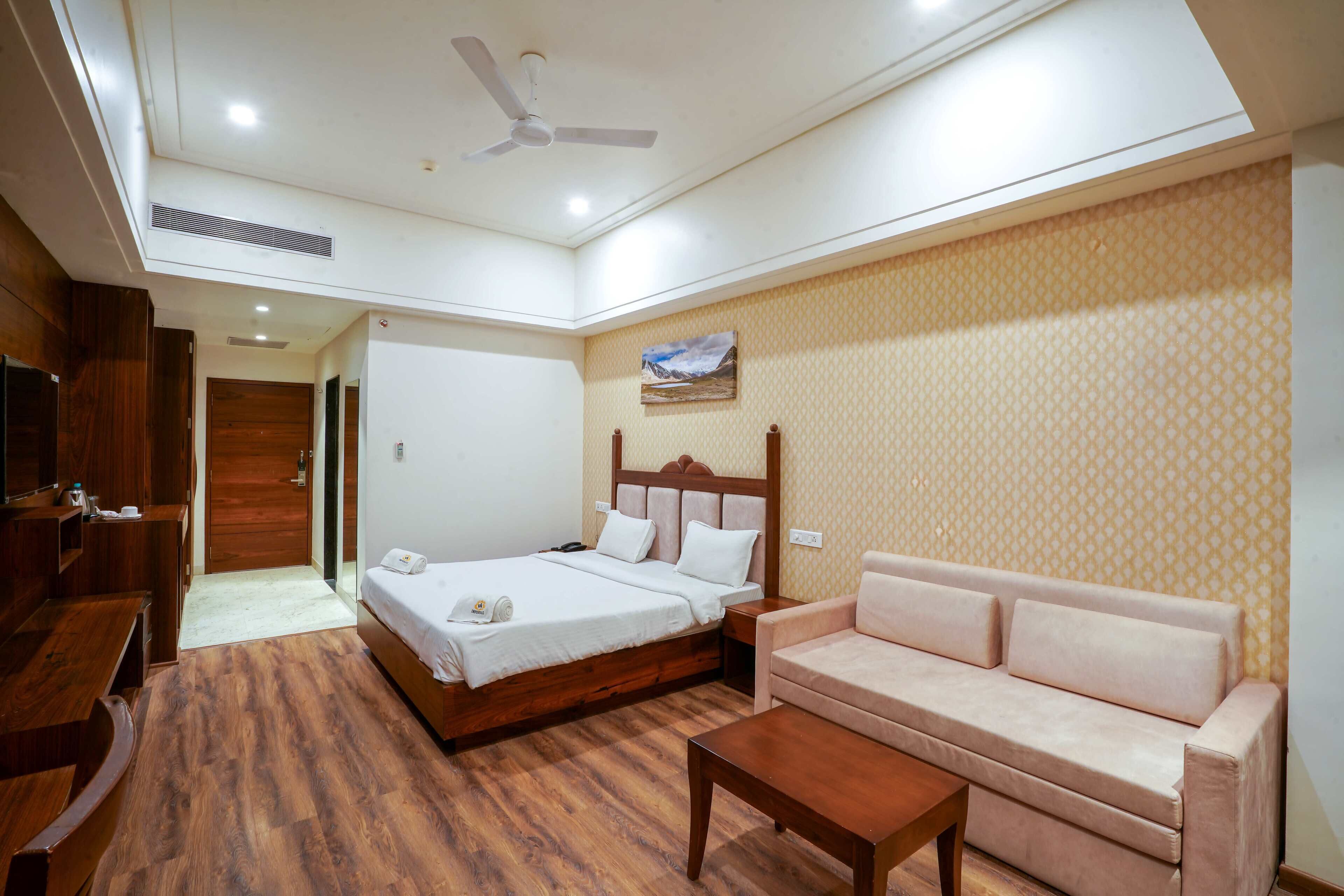Quarto de hotel com piso de madeira, cama com roupa de cama branca, sofá, mesa de madeira e ventilador de teto.