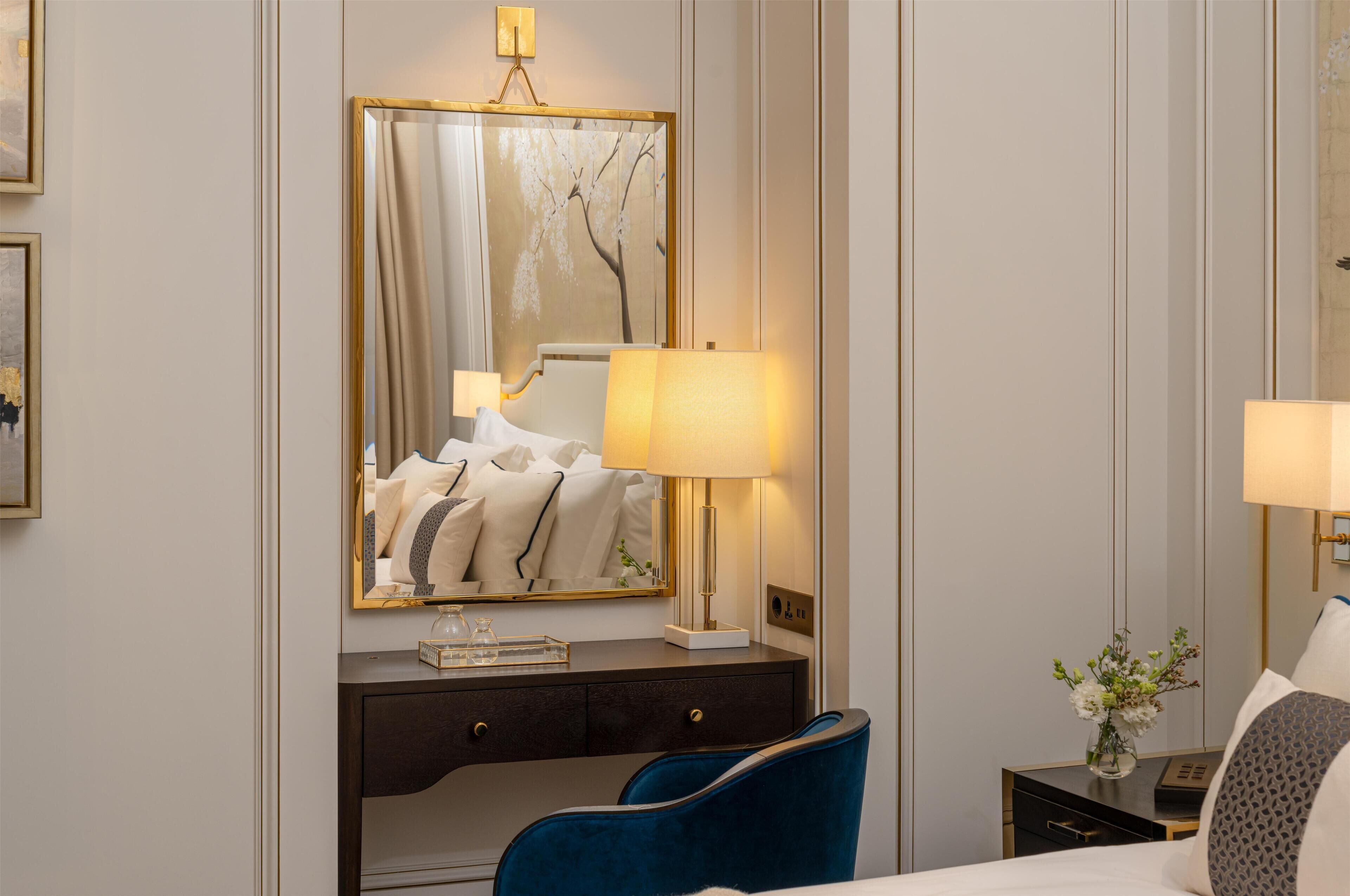 Quarto de hotel com um espelho grande, mesa de cabeceira com abajur, uma cadeira e uma pequena mesa com um vaso de flores.