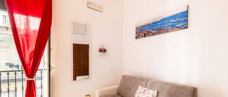 Uma sala de estar com sofá cinza, cortina vermelha, porta de madeira, interruptor de luz fixado na parede e um quadro com uma paisagem costeira.