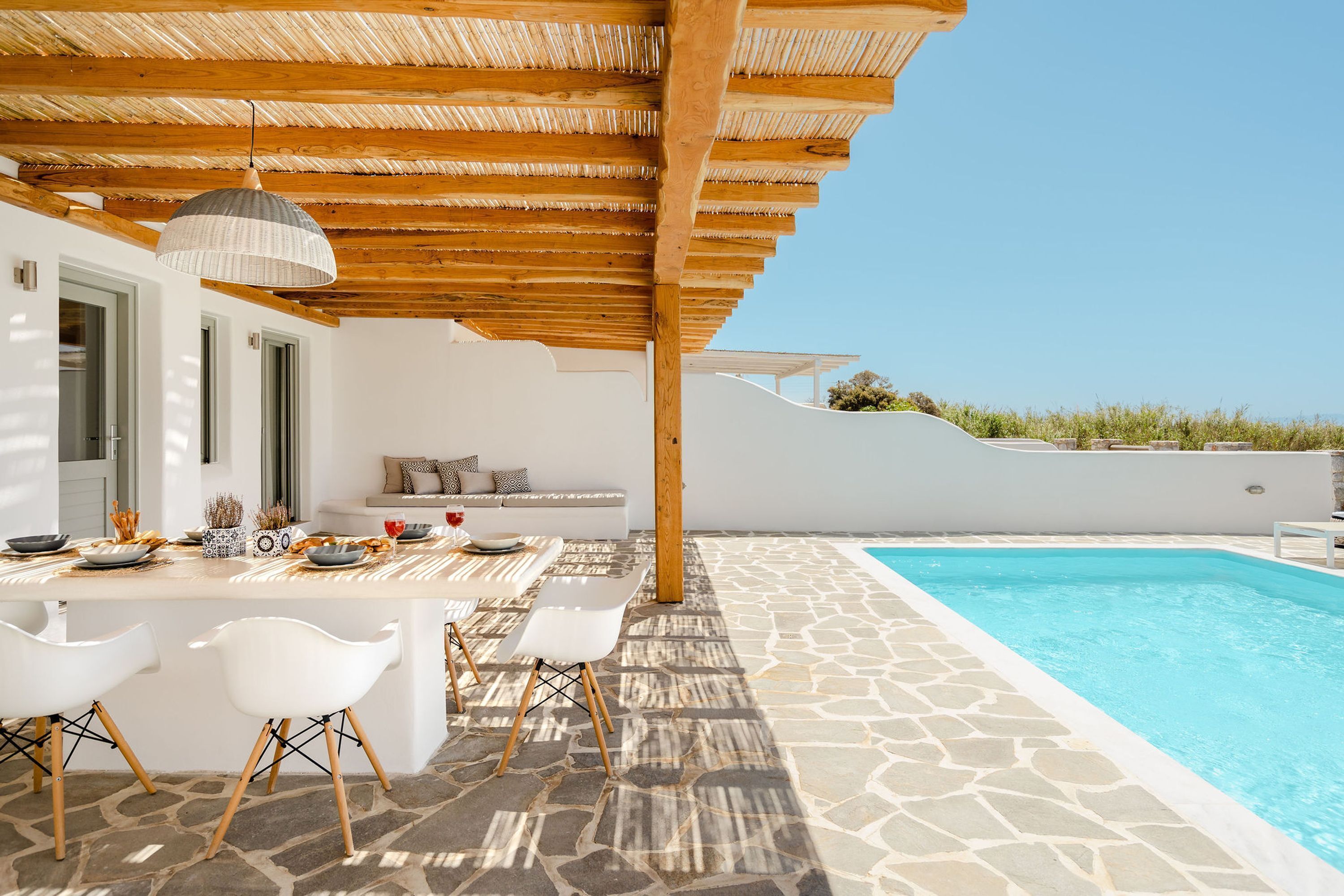 Uma área ao lado da piscina com um deck de pedra, uma pérgola de madeira e um espaço de refeições com cadeiras e mesa brancas.