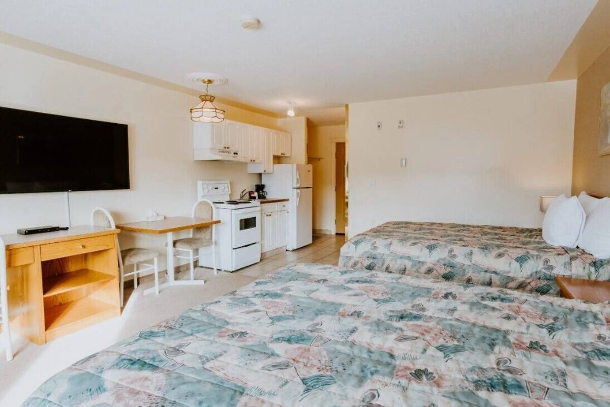 Quarto compacto com cama, escrivaninha, cadeira, televisão e pequeno espaço para cozinha.