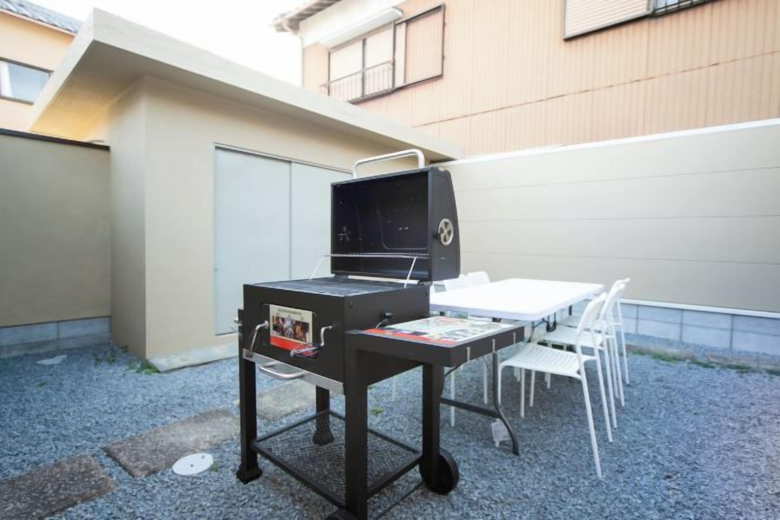 Área externa com churrasqueira, mesa e cadeiras, churrasqueira em carrinho e um edifício ao fundo.