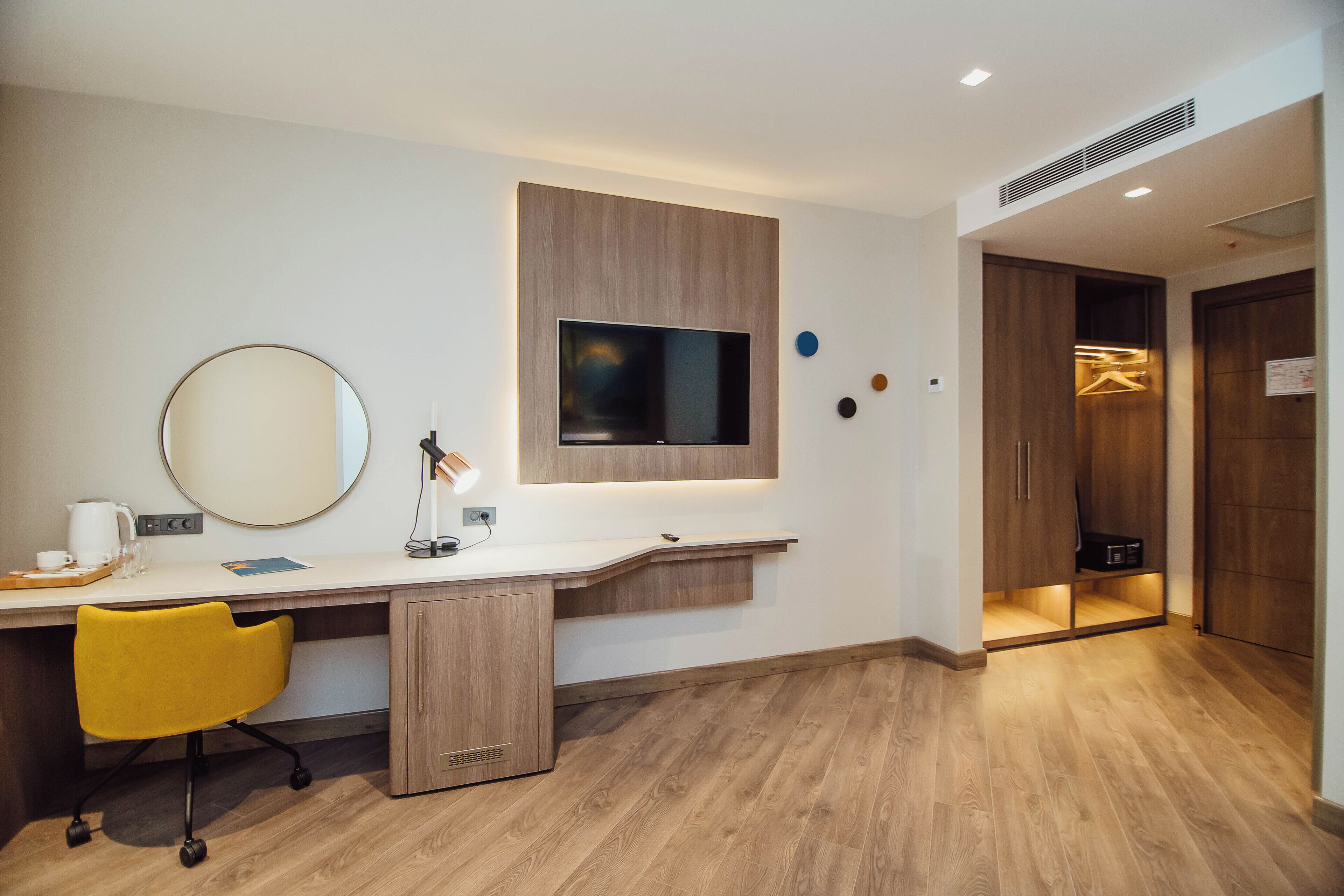 Quarto de hotel moderno com escrivaninha de madeira, espelho redondo, TV de tela plana e uma cadeira amarela.