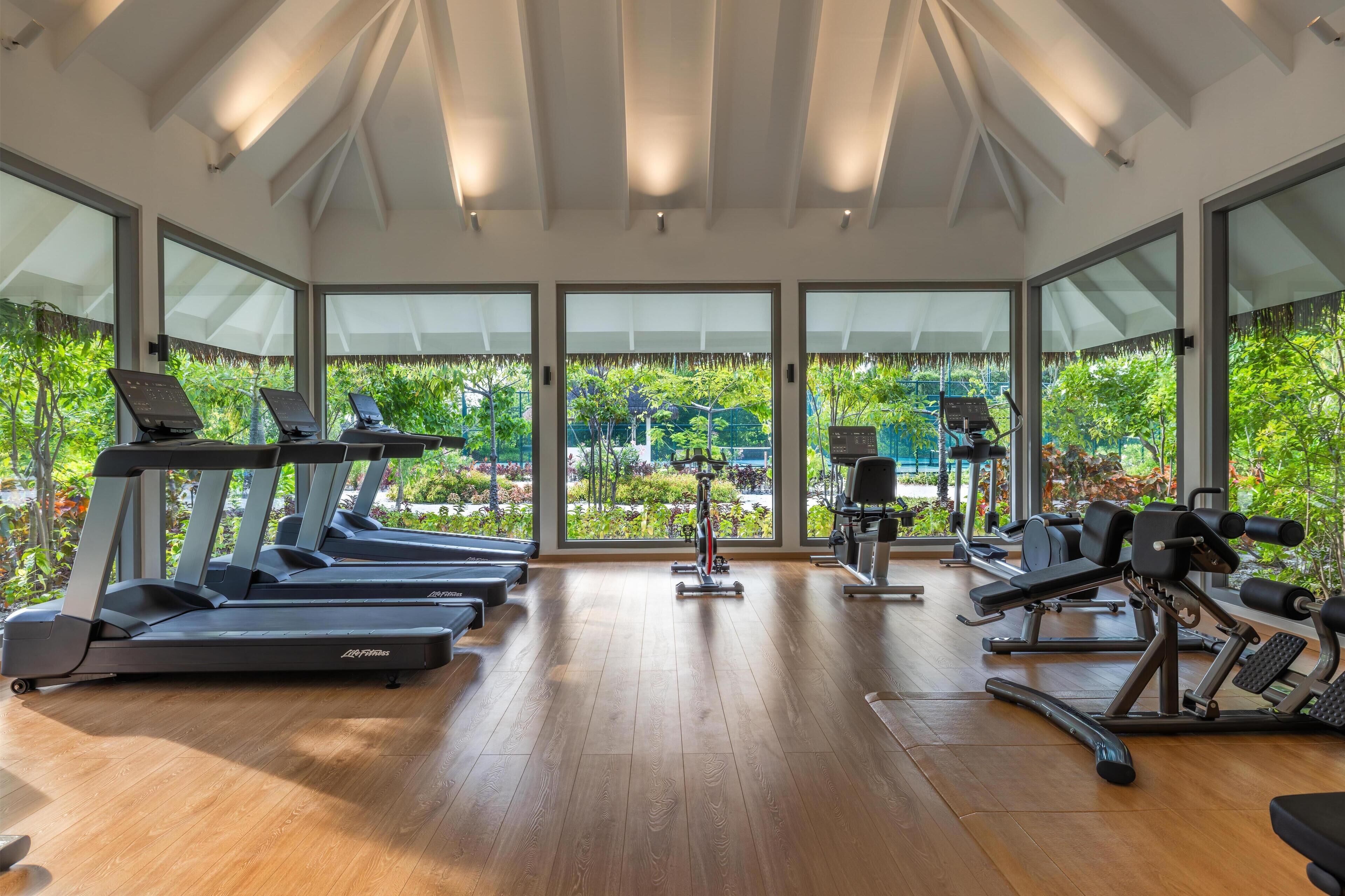 Academia com esteiras, elíptica e aparelhos de musculação. Grandes janelas proporcionam vista para o verde.