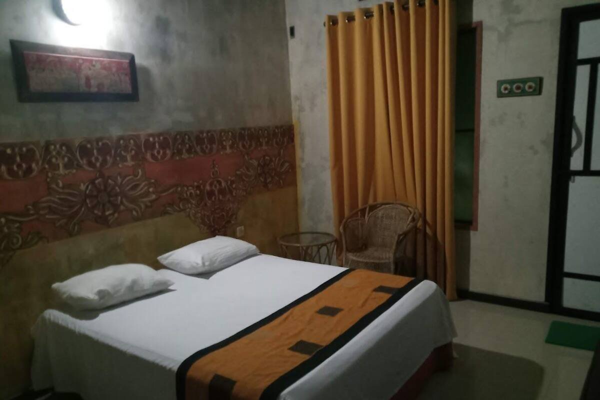 Uma cama com cabeceira estampada, uma cadeira e uma cortina.