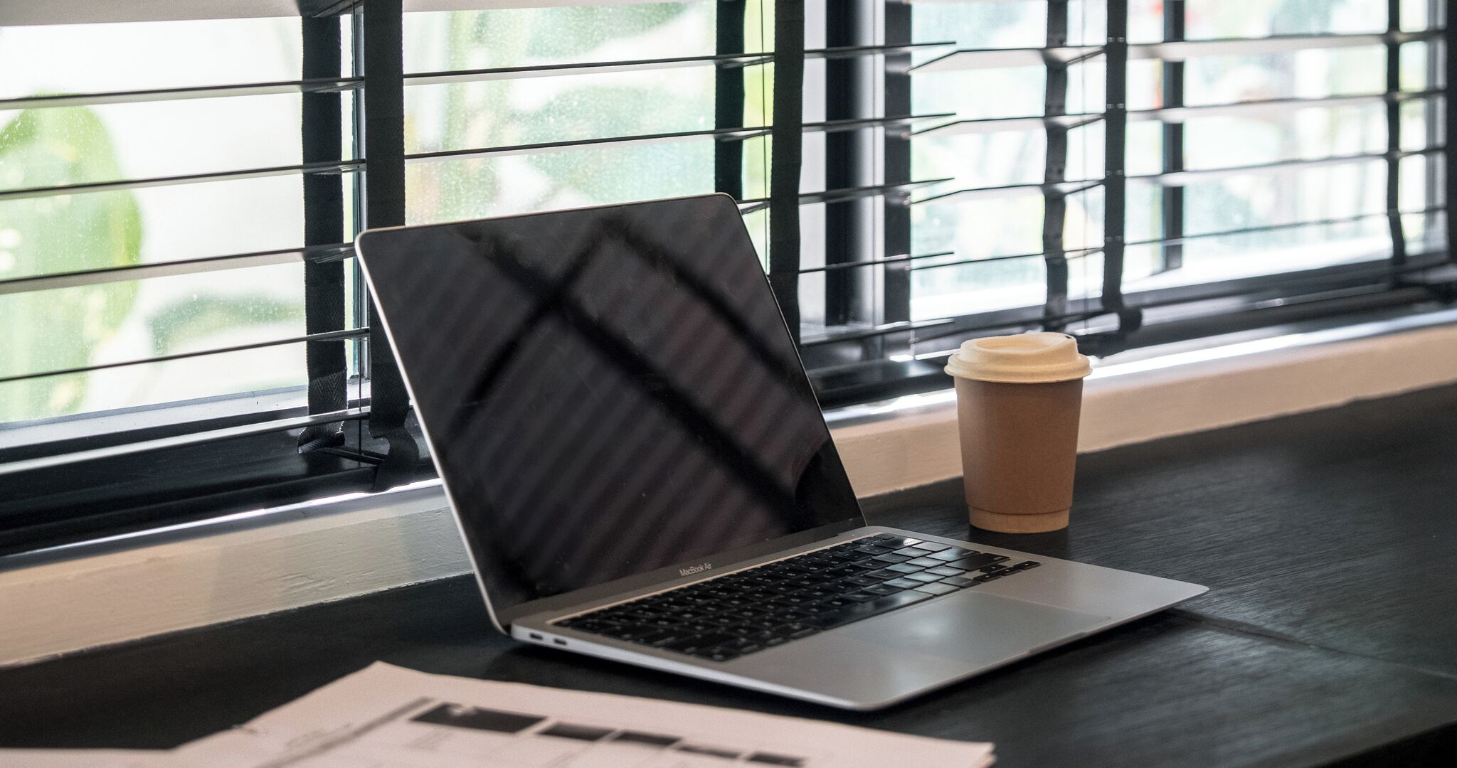 Um laptop sobre a mesa, com uma chávena de café por perto, perto de uma janela com persianas.