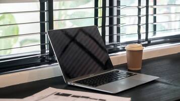 Um laptop sobre a mesa, com uma chávena de café por perto, perto de uma janela com persianas.