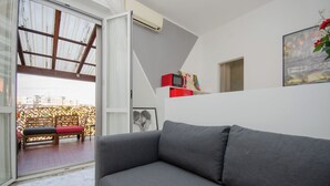 Uma sala de estar moderna com um sofá cinzento, um micro-ondas vermelho numa prateleira branca e uma varanda com um banco de madeira.