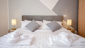 Cama bem arrumada com lençóis brancos e duas almofadas, duas luminárias de cabeceira e duas toalhas dobradas sobre a cama.