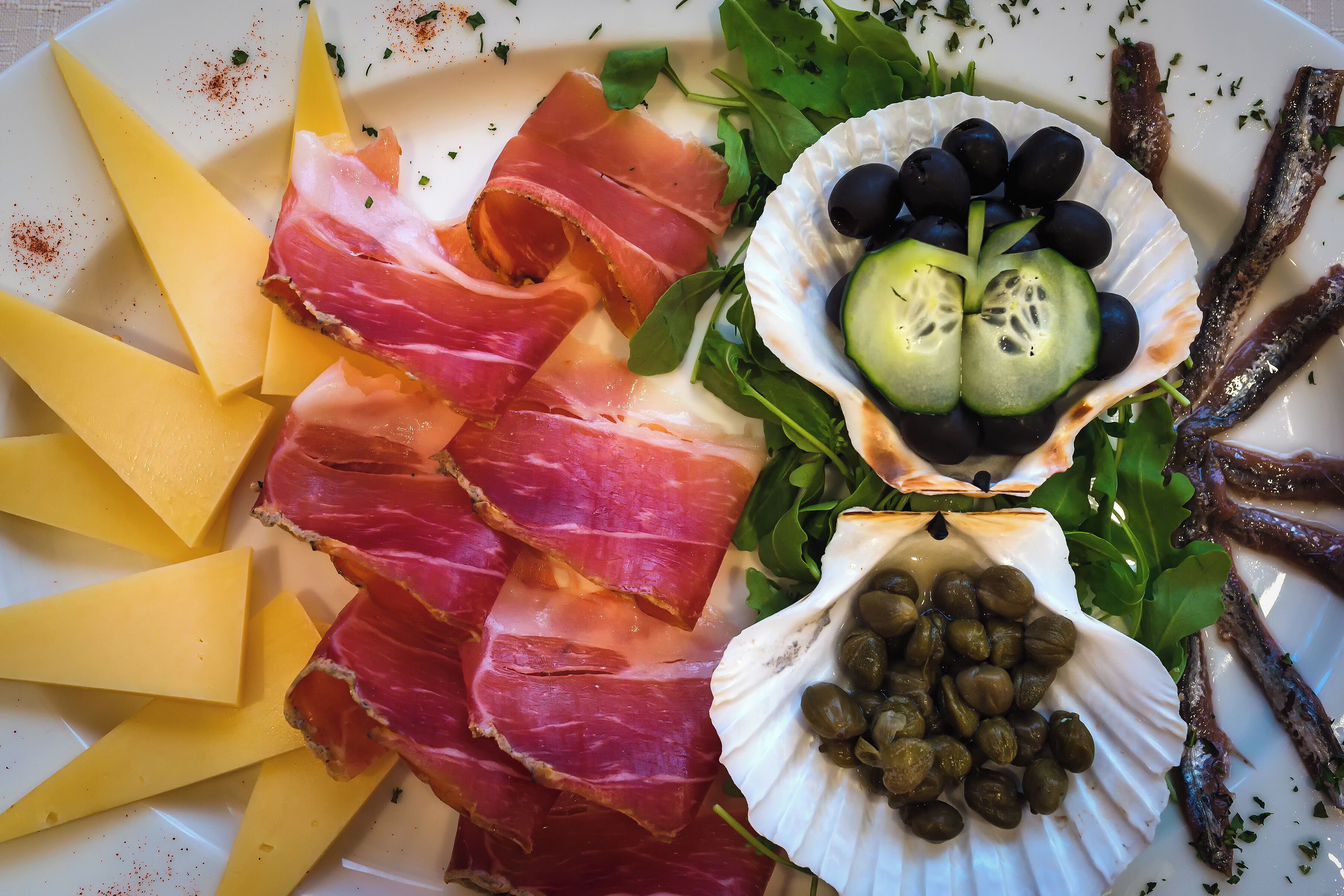 Um prato com diversos alimentos, incluindo presunto, queijo, azeitonas, alcaparras e vegetais.