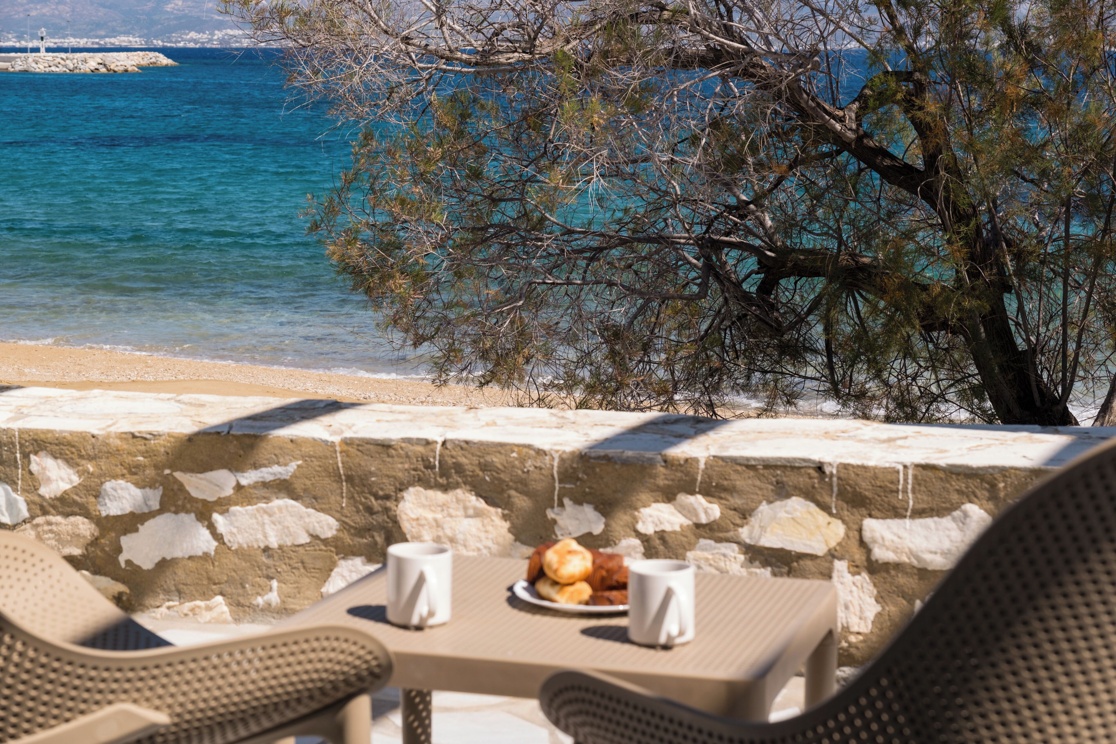 Uma mesa com duas canecas brancas e um prato de comida, com vista para uma praia e o mar.