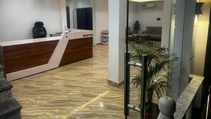 Uma área de recepção com balcão de madeira, um espaço de estar com sofá e mesa de centro, e uma planta em vaso.