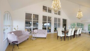 Uma sala de estar ampla, com teto alto, grandes janelas e um espaço de jantar com mesa e cadeiras de madeira.