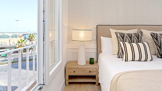 Quarto de hotel com uma janela ampla, mesa de cabeceira com abajur, cama com almofadas estampadas e vista para a paisagem urbana.