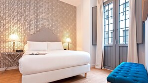 Quarto com cama, luminárias de cabeceira, um banquinho azul e parede estampada.