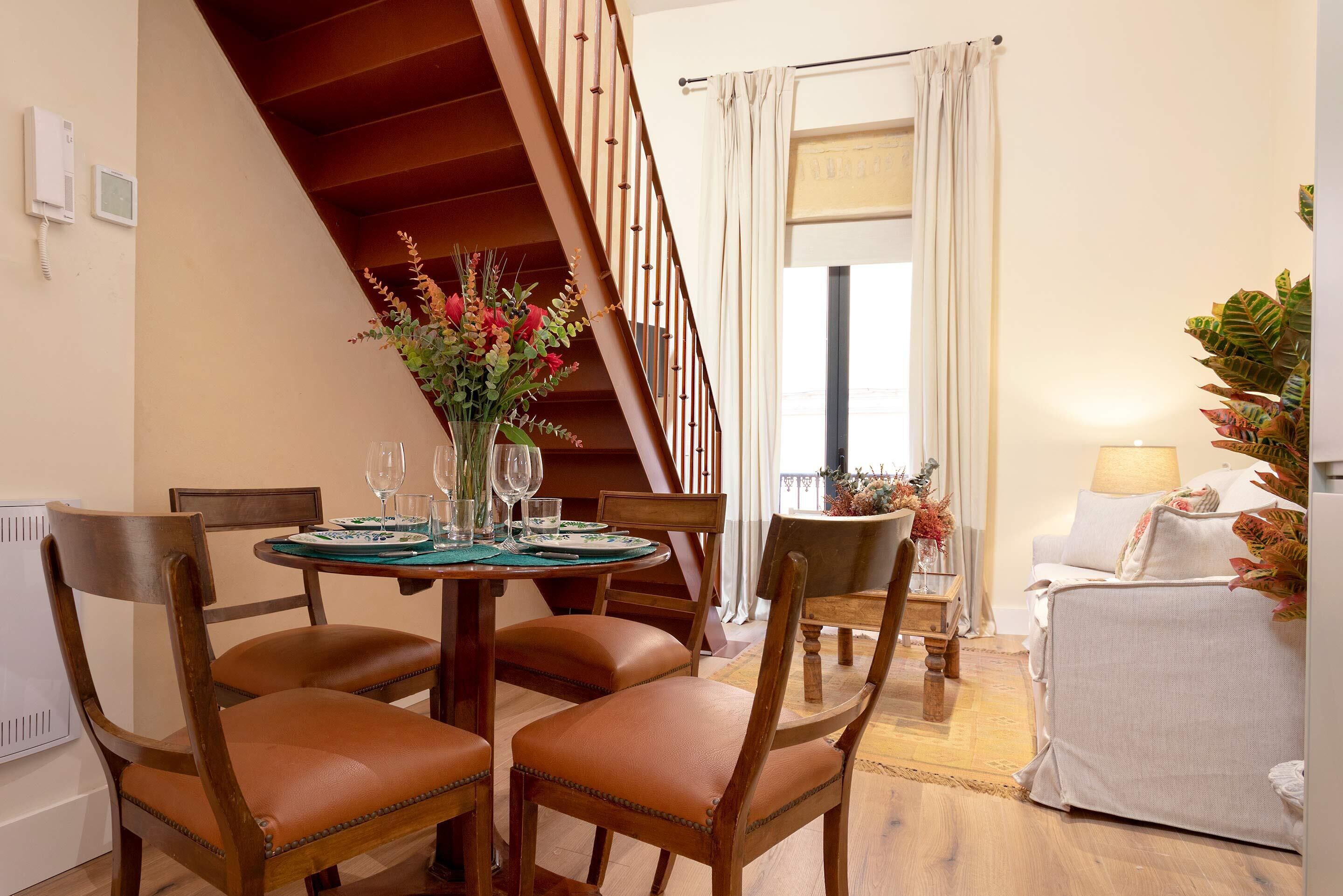 Uma sala de jantar com uma mesa de madeira, cadeiras e um vaso de flores. Uma escada é visível ao fundo.