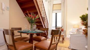 Uma sala de jantar com uma mesa de madeira, cadeiras e um vaso de flores. Uma escada é visível ao fundo.