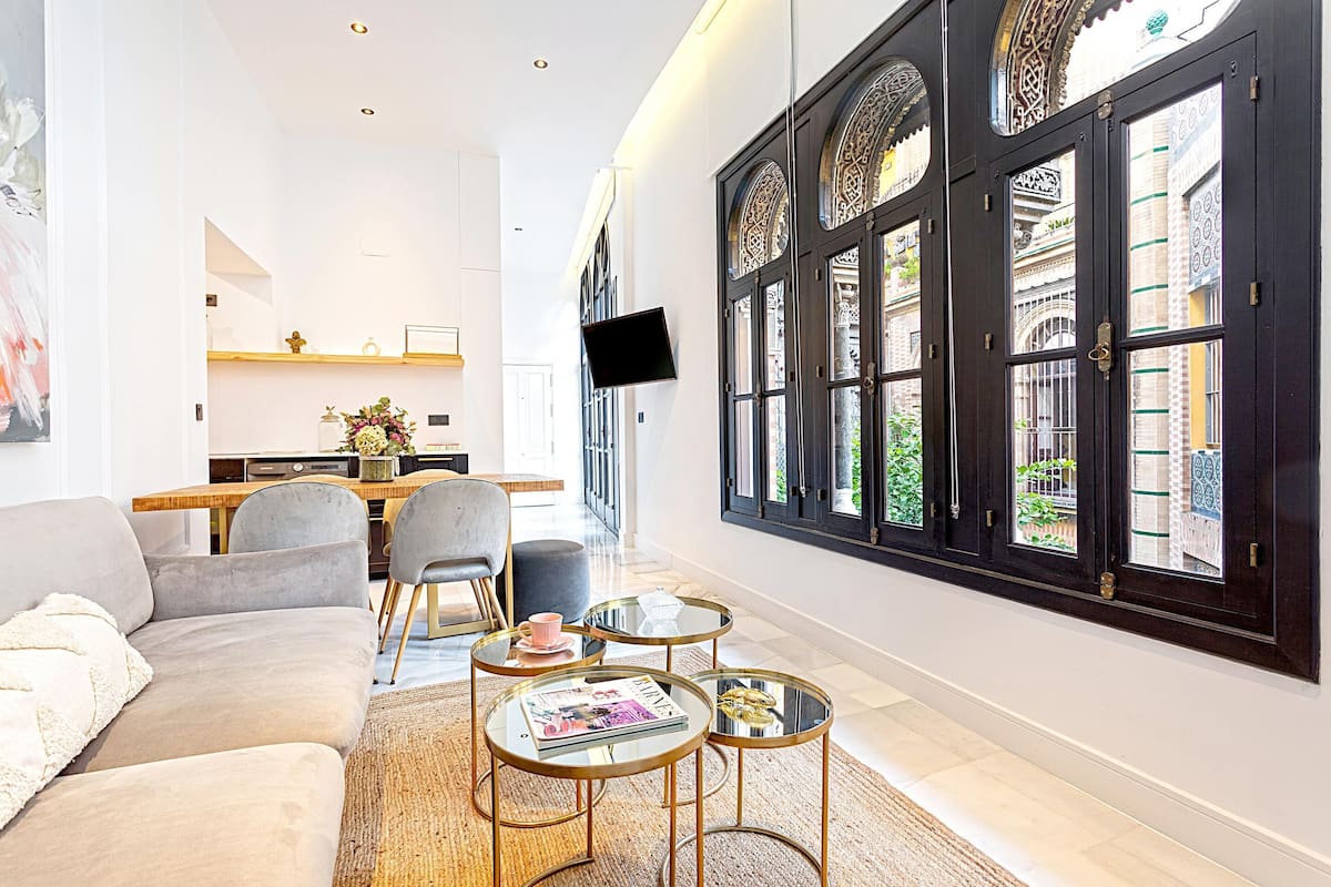Uma sala de estar moderna com sofá, mesa de centro e cadeiras, uma área de jantar com mesa e cadeiras, e janelas amplas com detalhe em ferro ornamental.