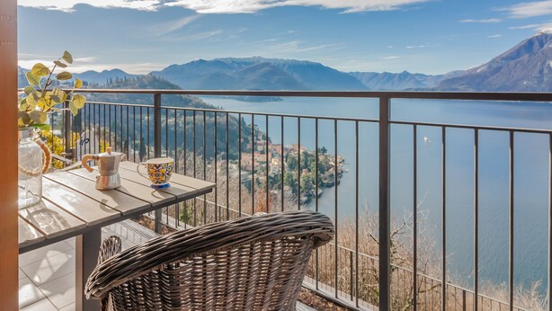 Varanda com vista para um lago e montanhas, uma cadeira de vime, uma mesa com uma xícara e um jarro, e um corrimão.