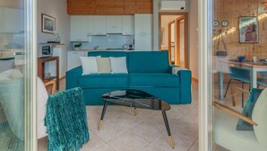 Uma sala de estar moderna com um sofá azul-turquesa, uma mesa de centro de vidro e uma cozinha ao fundo.