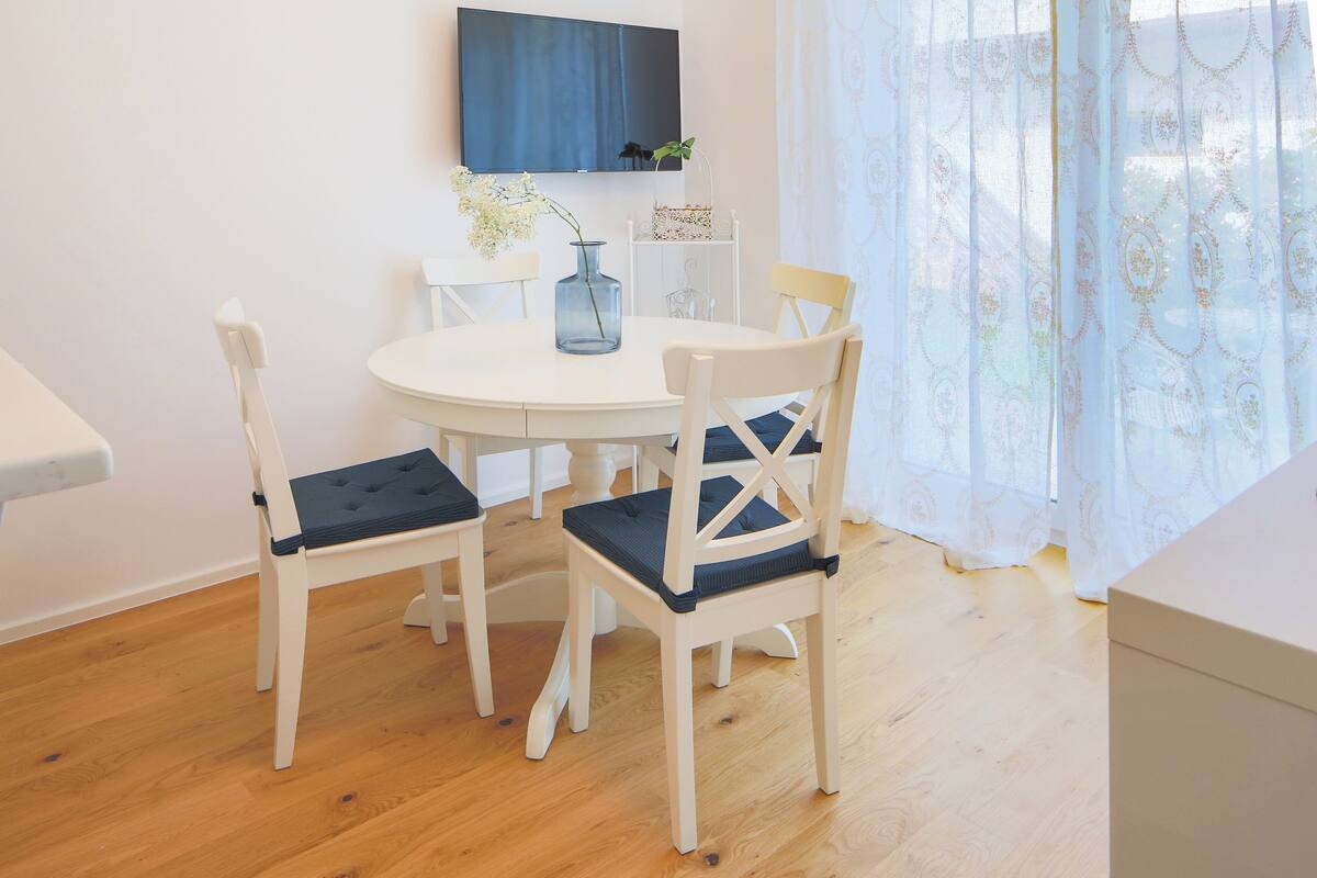 Uma área de refeições com uma mesa redonda, quatro cadeiras brancas com almofadas escuras, um vaso azul com flores, um armário branco e uma televisão montada na parede.