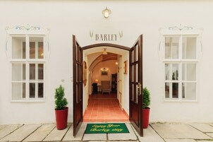 Entrada de hotel com uma placa indicando "BARLEY", um caminho de azulejos e duas plantas em vasos.