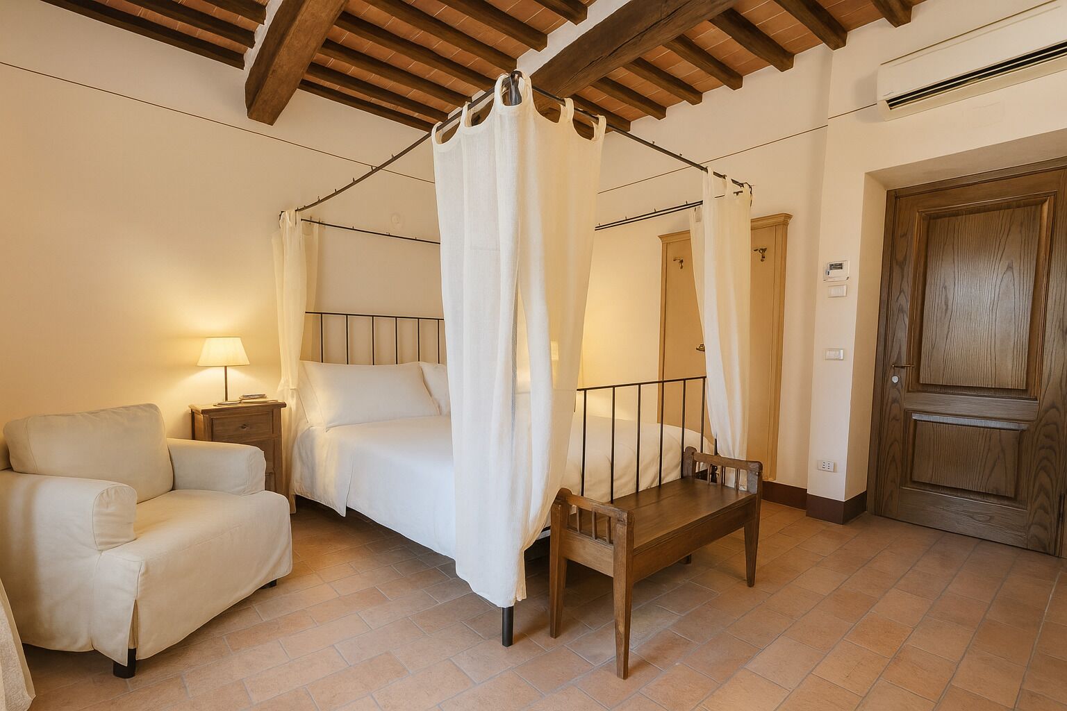 Quarto com cama de dossel, um banco de madeira, mesa de cabeceira com abajur, uma cadeira e porta de madeira.