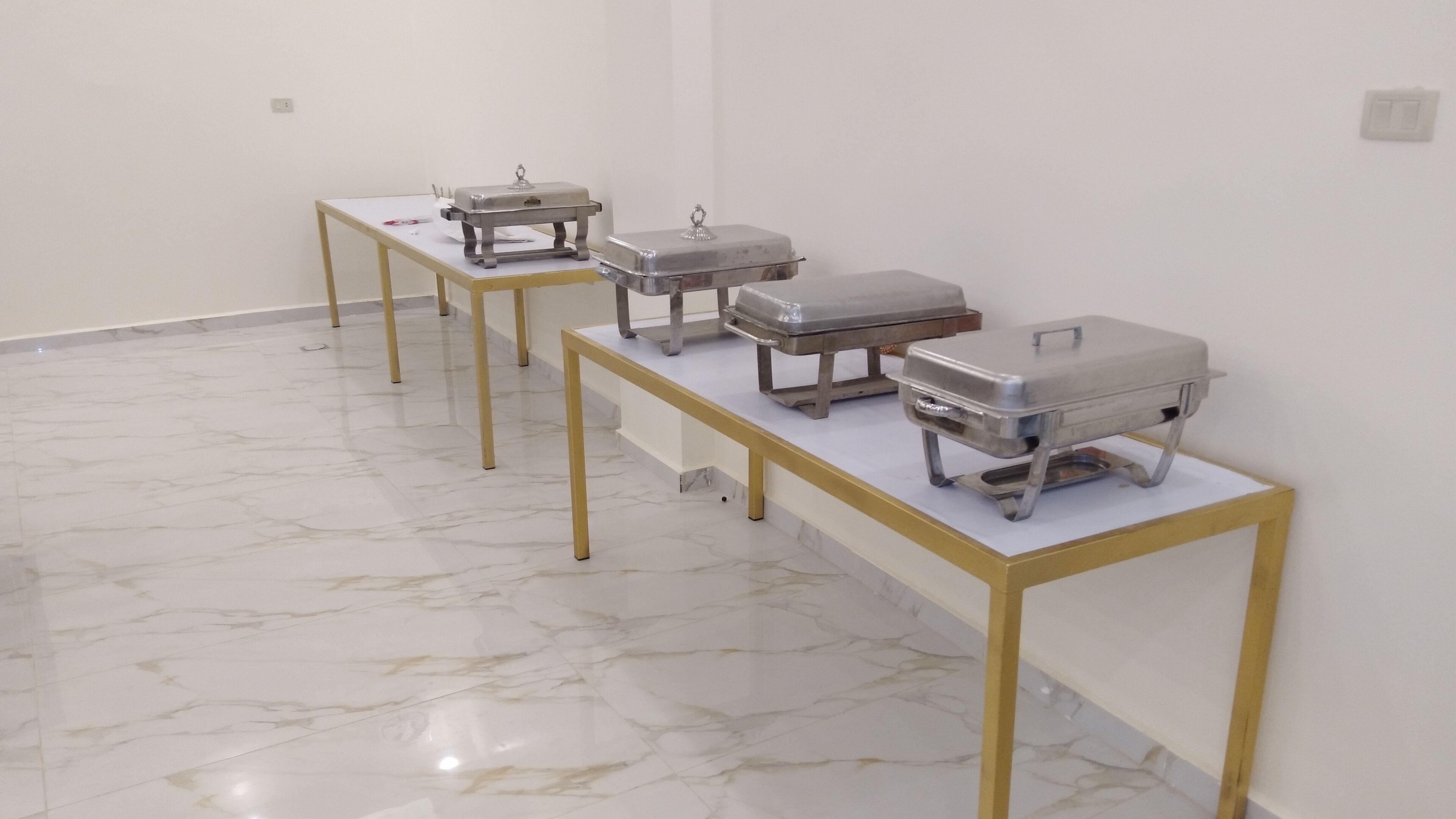 Uma fileira de mesas de buffet com rechauds de metal numa sala limpa e revestida a azulejos.