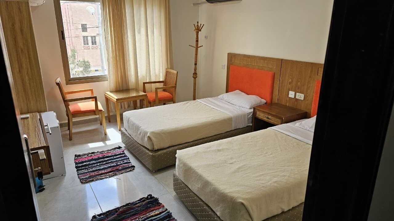 Quarto de hotel com duas camas, uma escrivaninha, uma cadeira e uma janela.