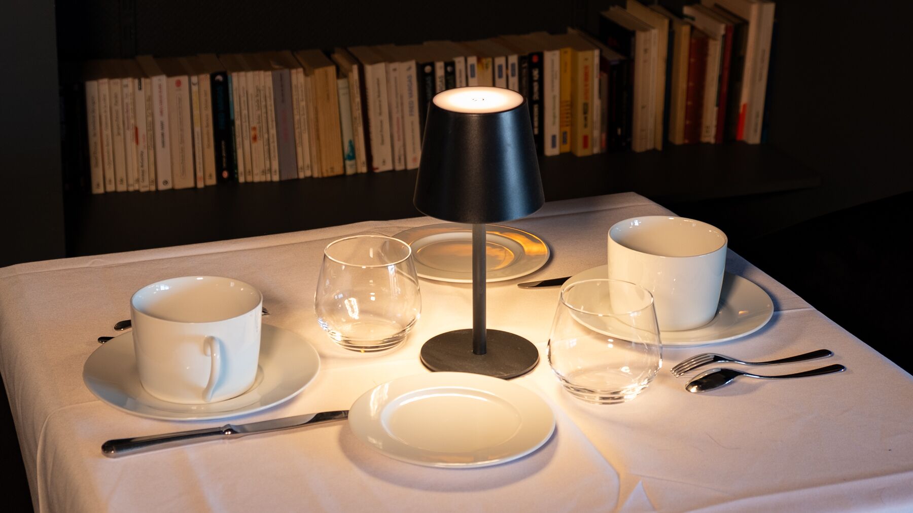Mesa posta para dois, com xícaras, pratos e talheres brancos, uma luminária preta e uma estante de livros ao fundo.