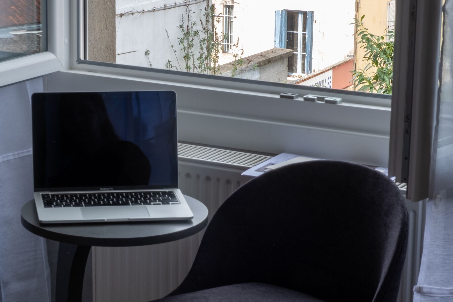 Um laptop sobre uma mesa pequena junto a uma janela, com um radiador ao fundo.