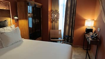 Quarto de hotel com cama, guarda-roupa, escrivaninha com luminária, cadeira e janela com cortinas.