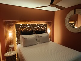 Cama bem arrumada com roupa de cama e almofadas brancas com a inscrição "DALMA" em um quarto com paredes marrons e cabeceira de madeira.