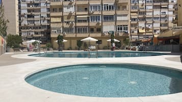 Piscina com um calçadão ao redor, um edifício com vários andares e varandas, e áreas de estar ao ar livre.