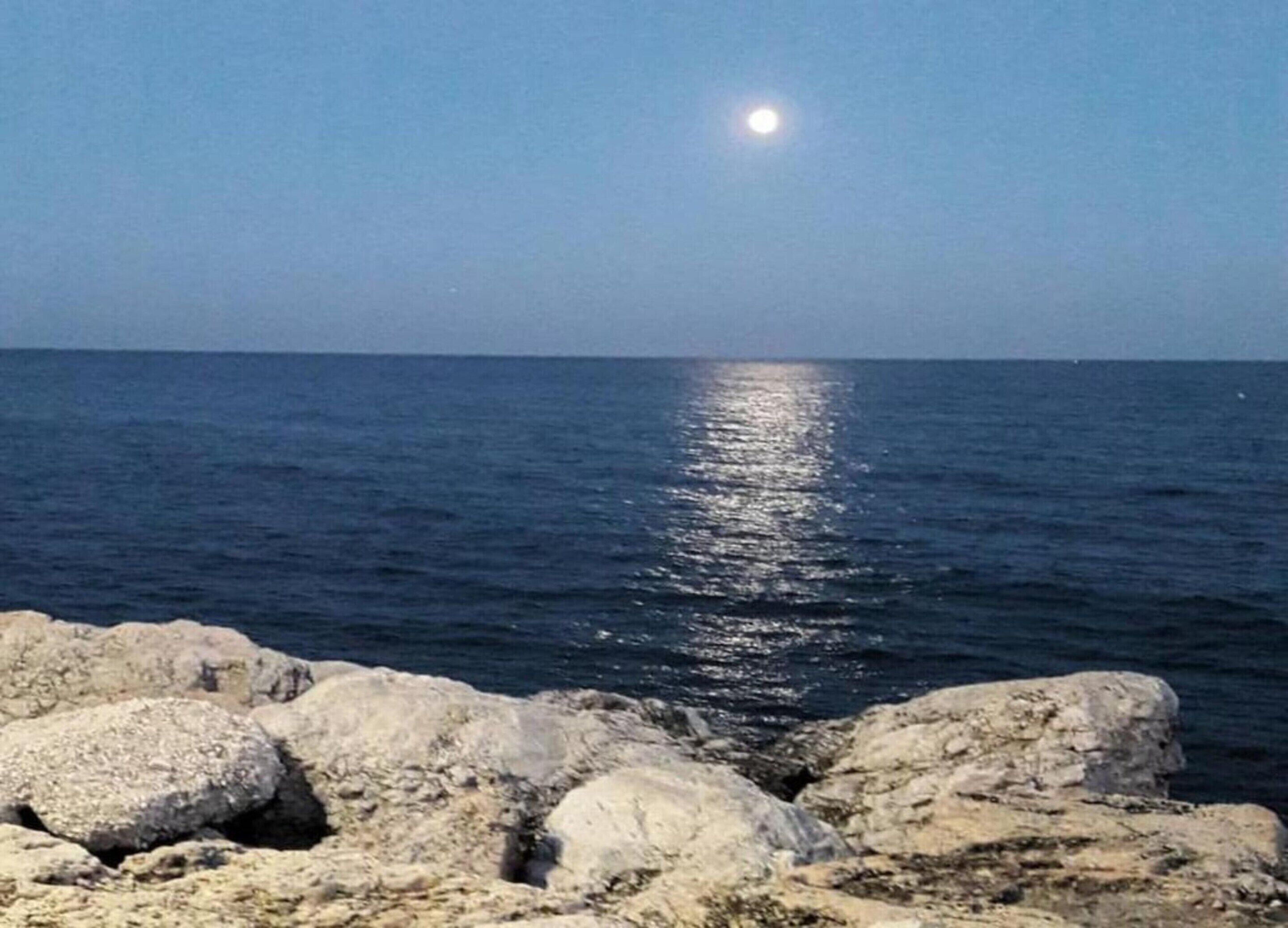 Costa rochosa com mar calmo e lua visível.
