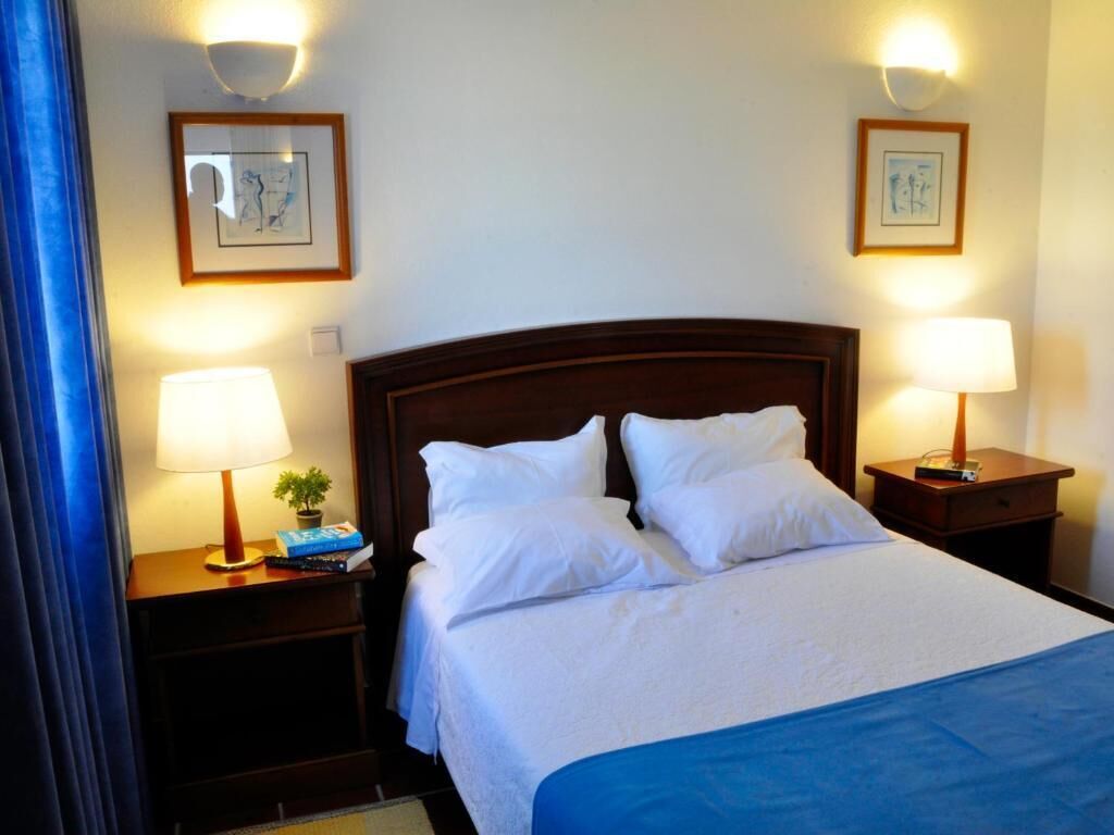 Cama bem arrumada com lençóis brancos, duas luminárias de cabeceira e obras de arte emolduradas nas paredes.
