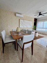 Uma sala de jantar moderna com uma mesa de madeira, cadeiras estofadas em cinza claro, um vaso grande e um ventilador de teto.