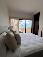 Quarto de hotel com cama, mesa de cabeceira, luminária e vista para uma varanda com guarda-corpo.
