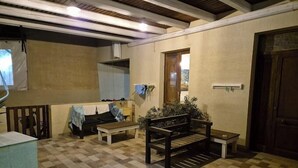 Uma sala de estar aconchegante com piso xadrez, um banco de madeira, um sofá e uma pequena mesa. Há uma porta à direita e uma janela à esquerda.