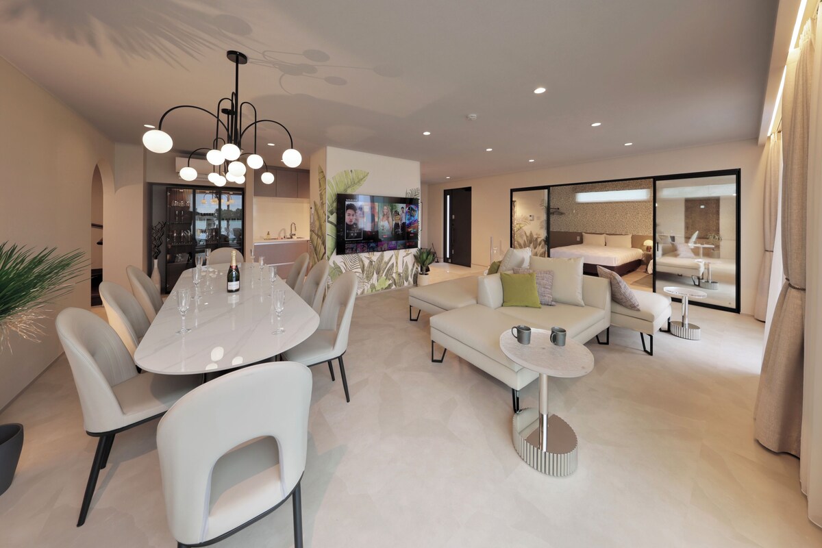 Quarto de hotel moderno com área de jantar, sofá, televisão e varanda.